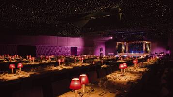 Salão de festas com mesas montadas para um evento formal, com luminárias vermelhas e um palco ao fundo.