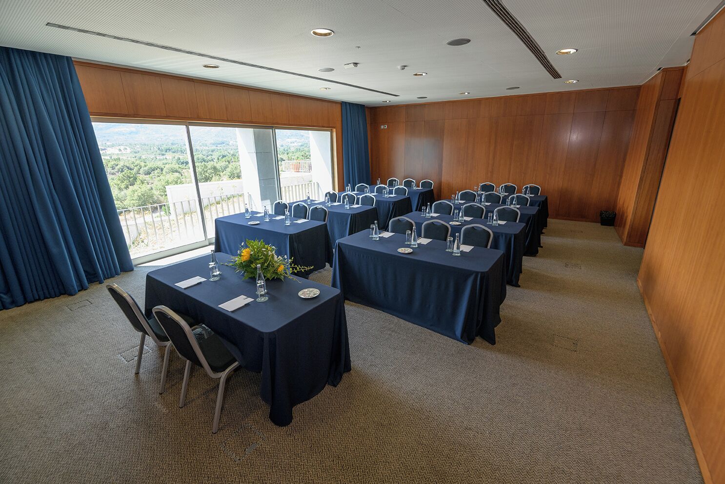 Uma sala de conferências com várias mesas, cadeiras e uma grande janela que oferece vista para o verde.