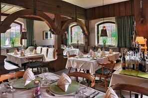 Um restaurante com vigas de madeira, mesas redondas com toalhas brancas e cadeiras verdes.