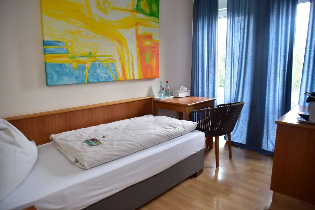 Quarto de hotel com uma cama, escrivaninha, cadeira e um quadro abstrato colorido na parede.