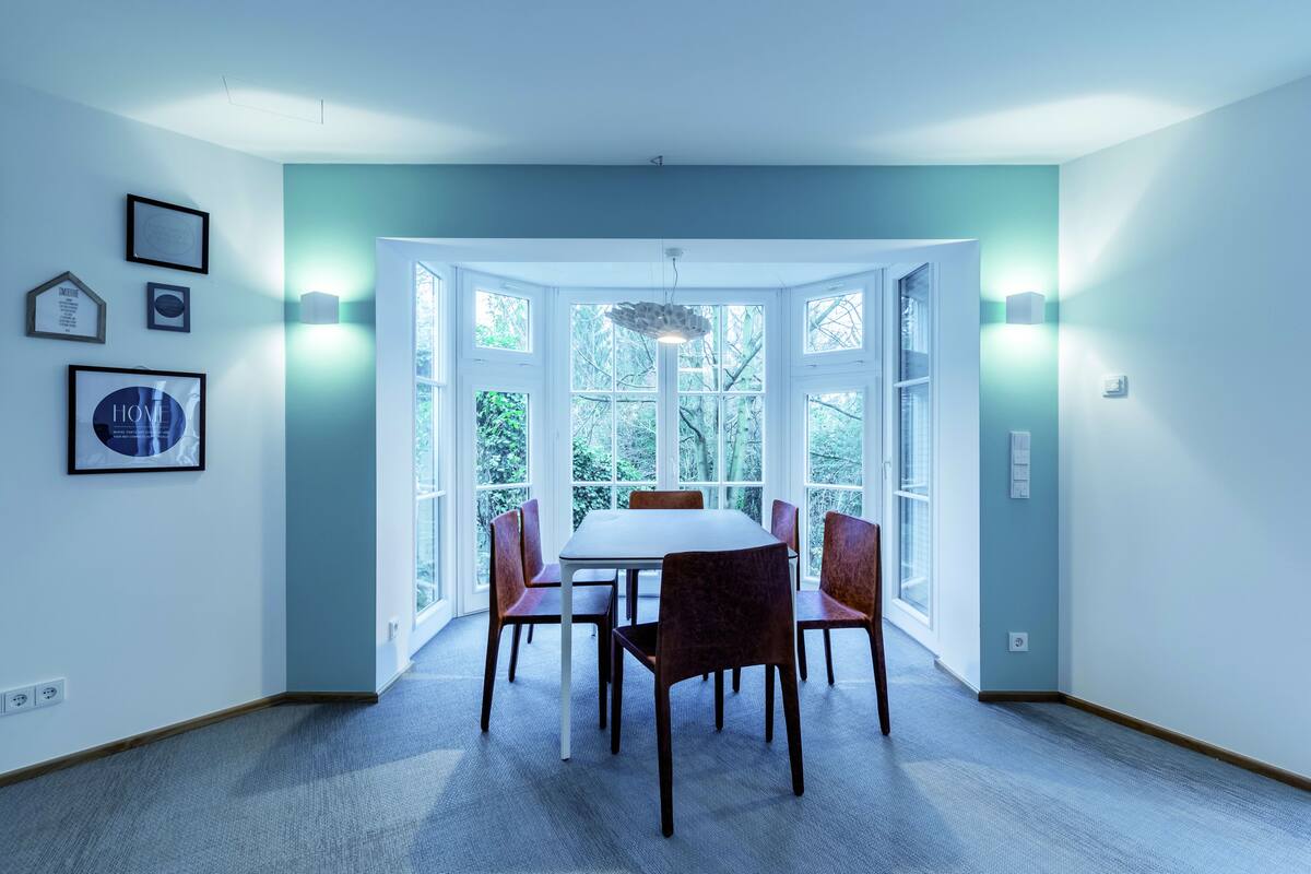 Uma área de jantar com mesa e cadeiras, janelas amplas e luminárias fixadas na parede.