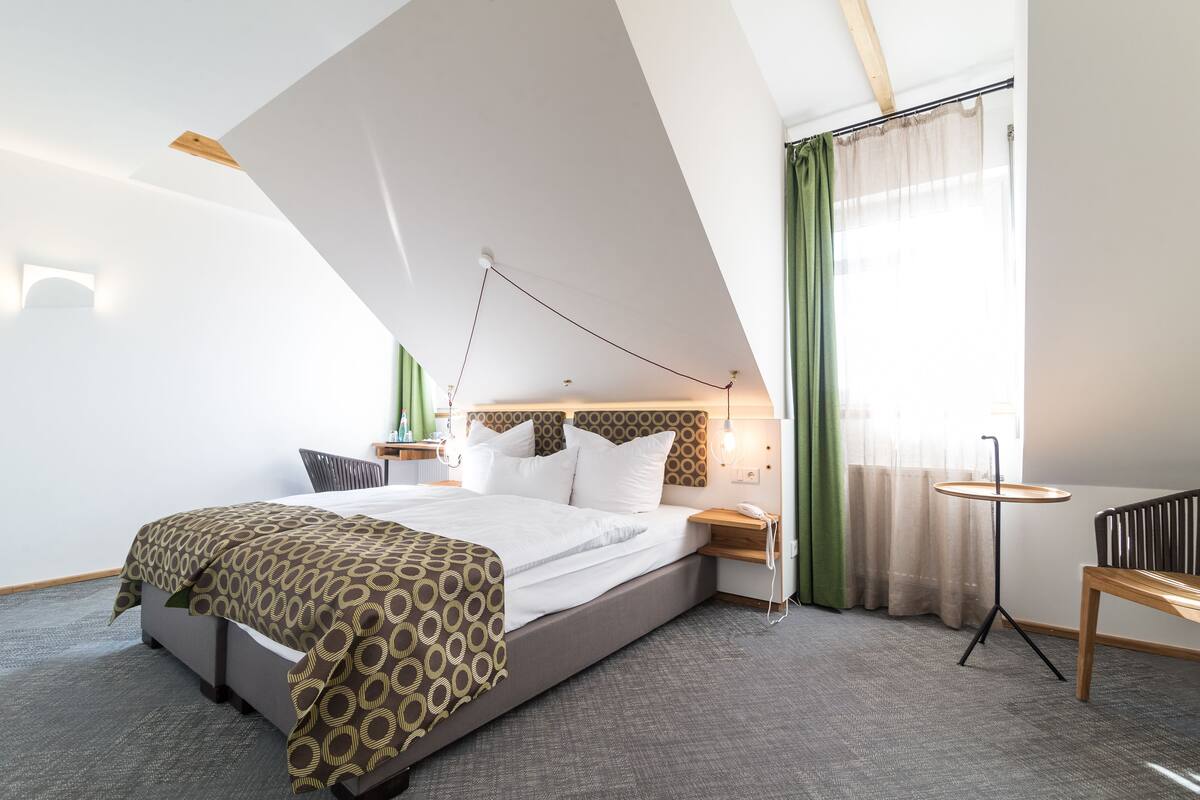 Quarto de hotel moderno com uma cama grande, uma pequena mesa redonda, uma cadeira e uma mesa de cabeceira com abajur.