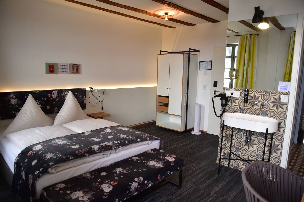 Quarto de hotel com cama, mesa de cabeceira, espelho, pia e área de chuveiro.