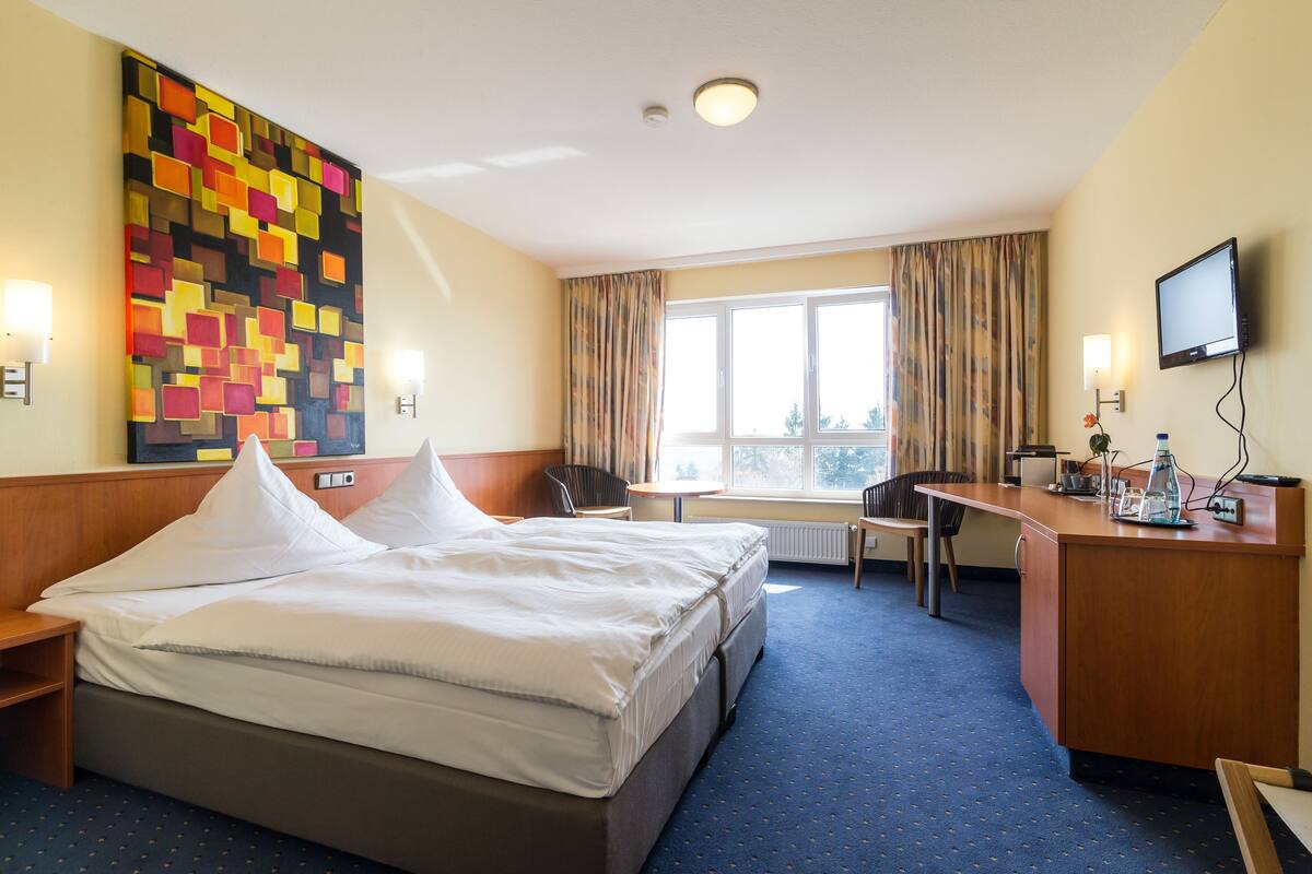Quarto de hotel com cama grande, escrivaninha, cadeira, televisão e janela com cortinas.