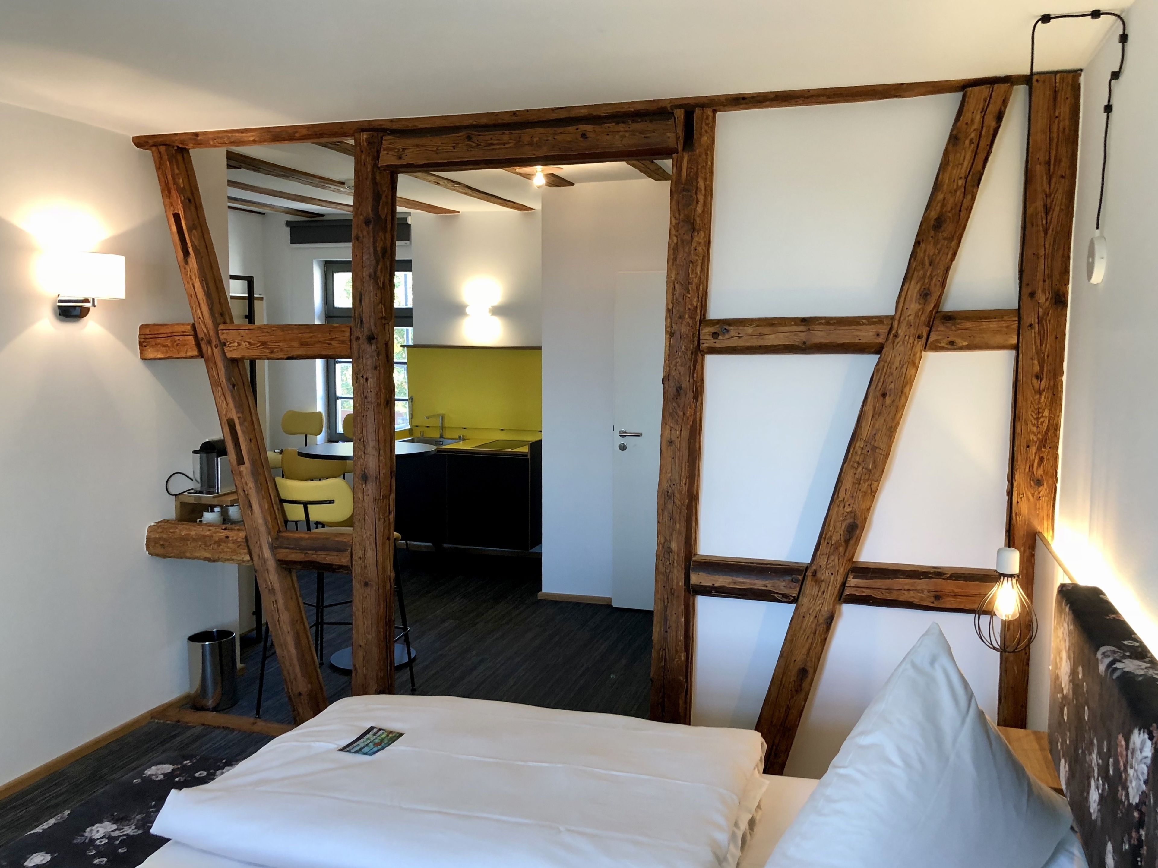 Quarto com cama, vigas de madeira, uma cadeira amarela, uma mesa pequena e área de cozinha ao fundo.