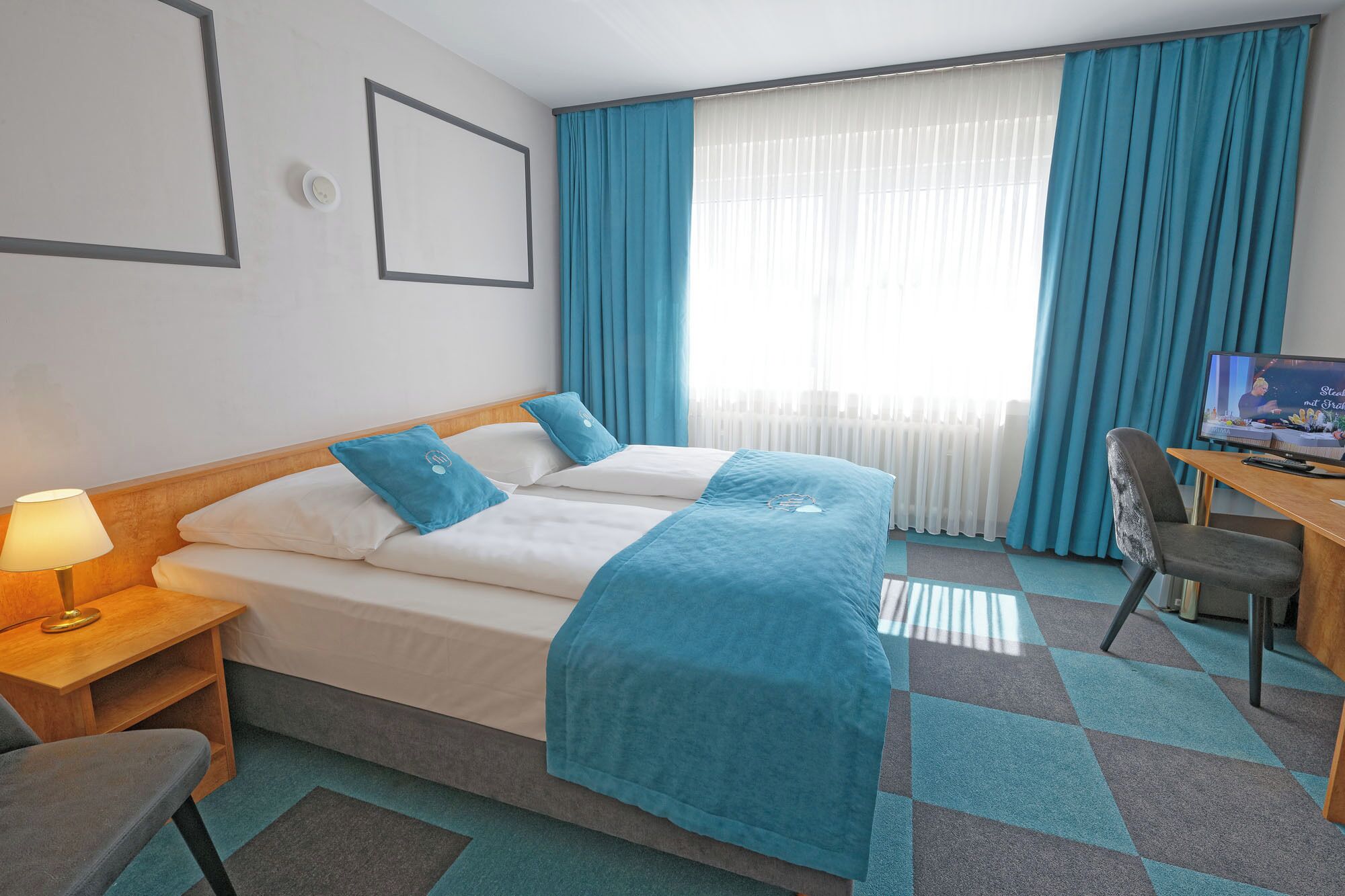Quarto de hotel com cama, escrivaninha, cadeira, televisão e janela com cortinas.