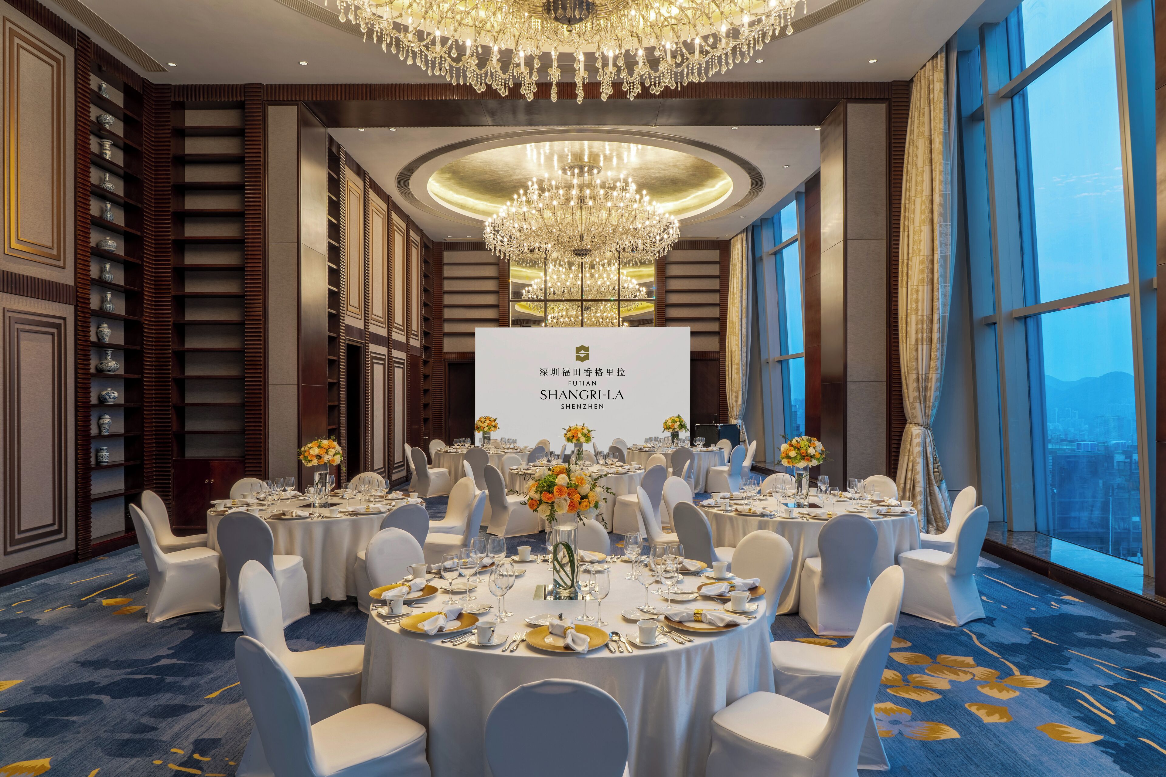 Uma sala de banquetes com mesas redondas preparadas para um evento formal, um grande candelabro e uma faixa com o nome do hotel.