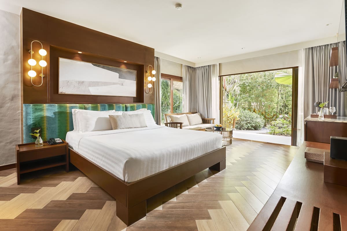 Quarto de hotel moderno com uma cama grande, uma mesa de cabeceira de madeira, um espaço de estar com uma mesa e vista para o verde lá fora.