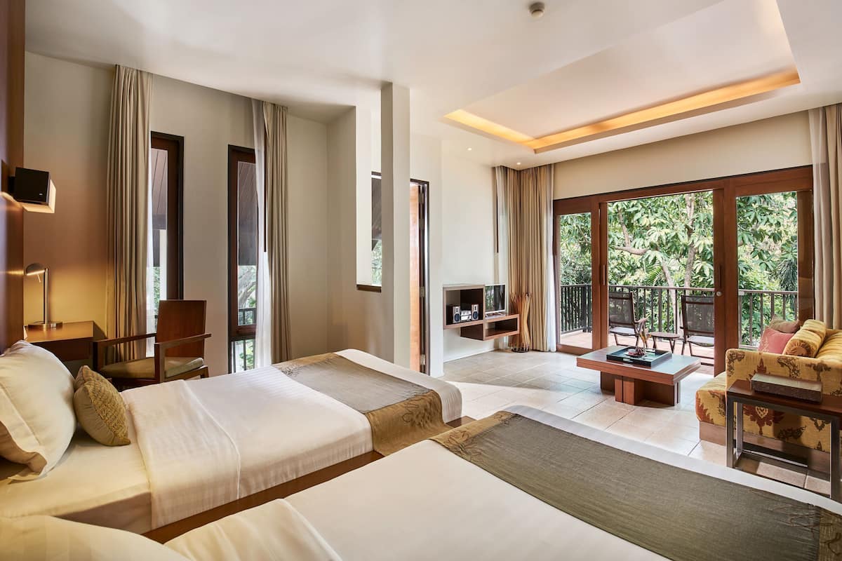 Quarto de hotel com duas camas, um sofá, uma TV e uma varanda com vista para o verde.