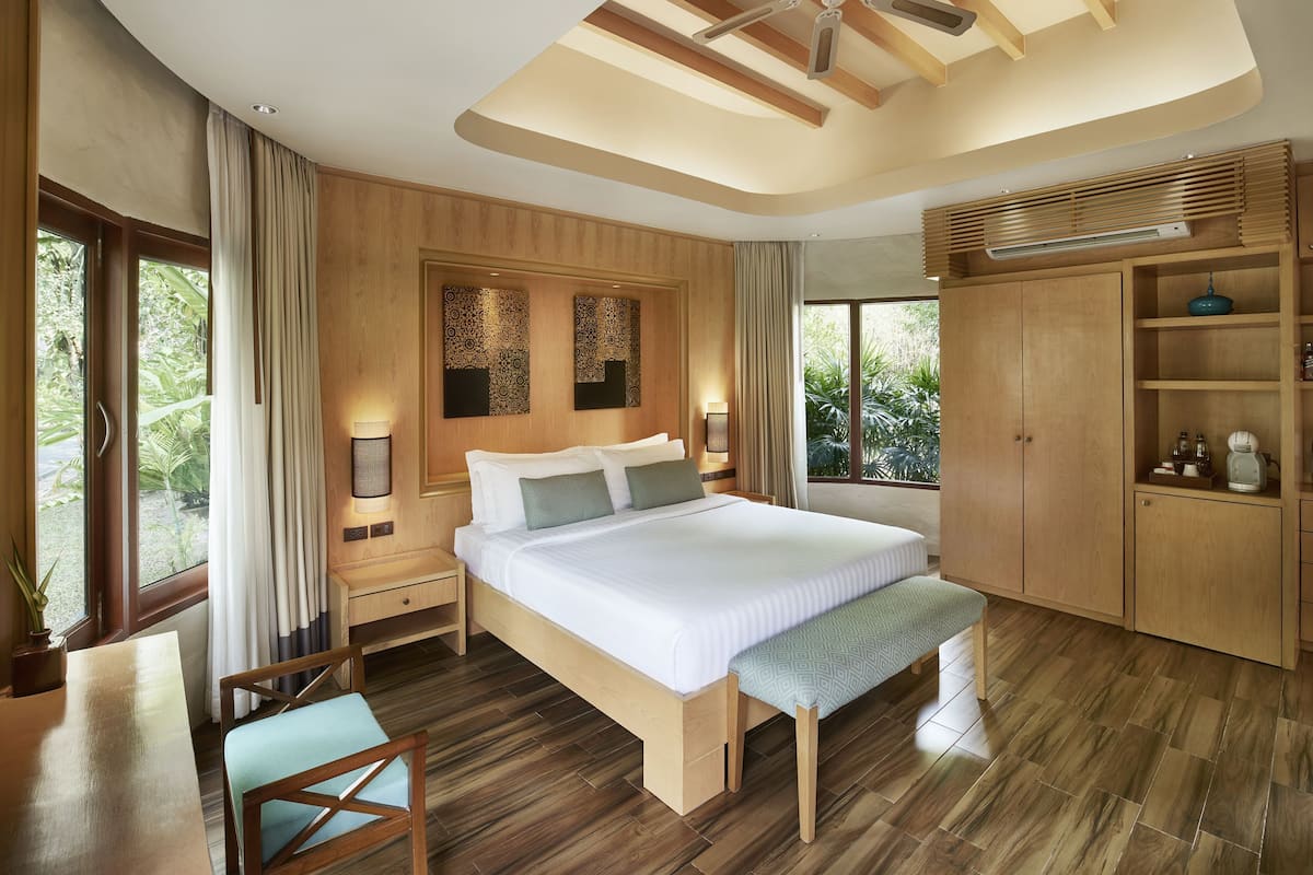 Quarto moderno com cama grande, piso de madeira, ventilador de teto e vista para área verde.