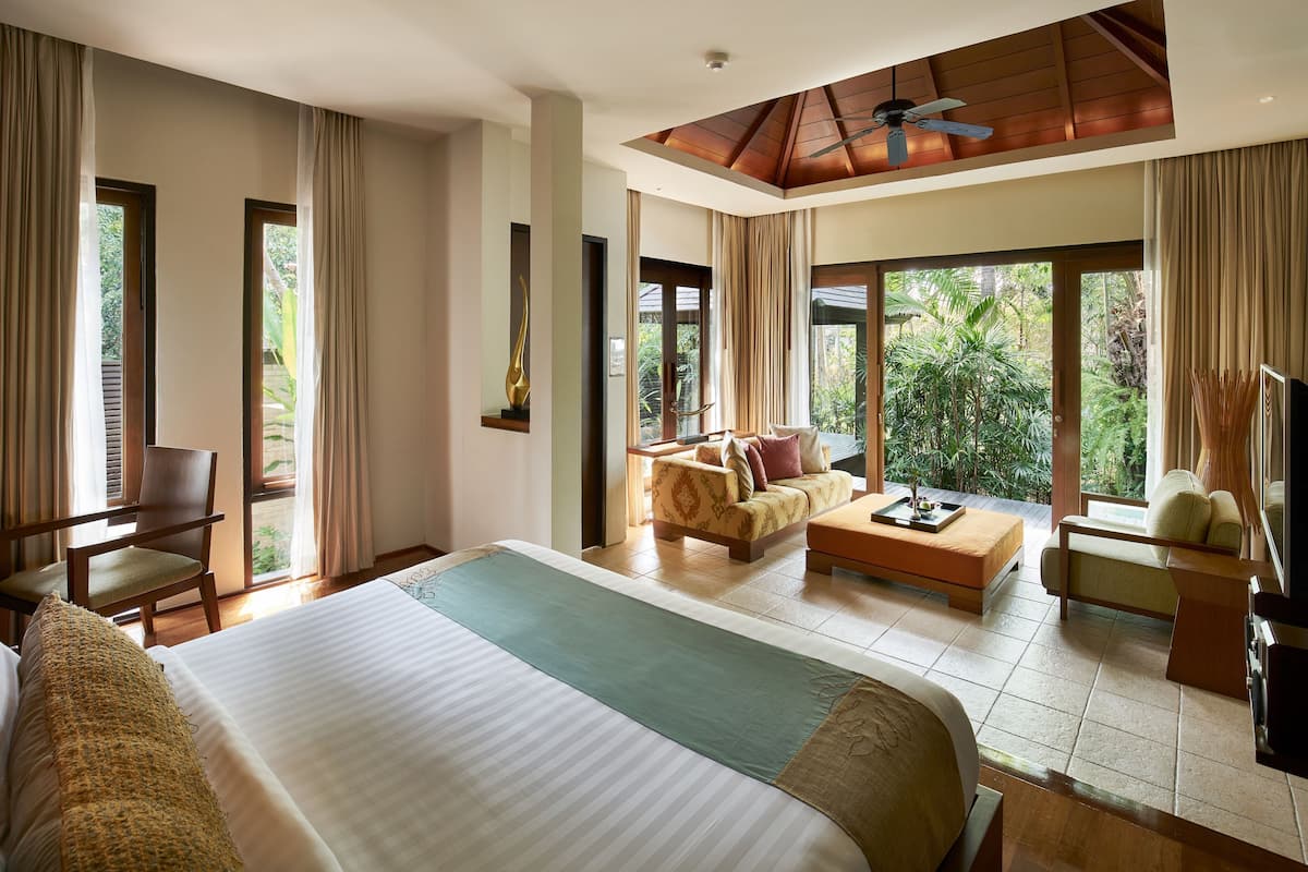 Quarto de hotel espaçoso com uma cama grande, um sofá, uma cadeira e uma mesa de trabalho. Amplas janelas oferecem vista para o exterior.