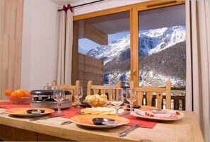 Mesa de jantar posta com pratos, copos e uma cesta de laranjas, com vista para uma montanha pela janela.