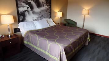 Quarto de hotel com cama, duas luminárias de apoio, uma mesa de cabeceira, uma cadeira e um quadro grande pendurado na parede com uma cachoeira.