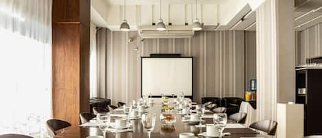 Uma sala de conferências com uma mesa comprida preparada para uma reunião, equipada com cadeiras, mesas e uma tela de projetor.