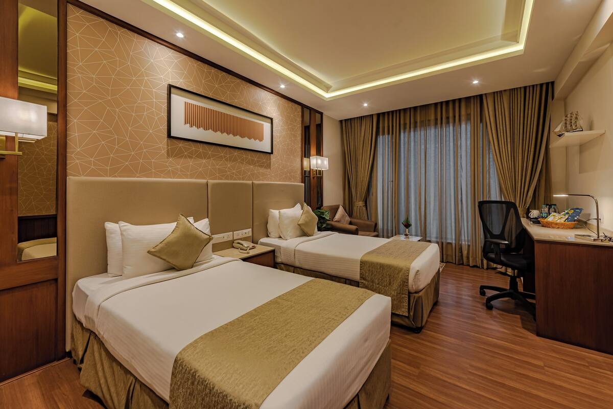 Quarto de hotel com duas camas, uma escrivaninha, uma cadeira e uma janela grande com cortinas.