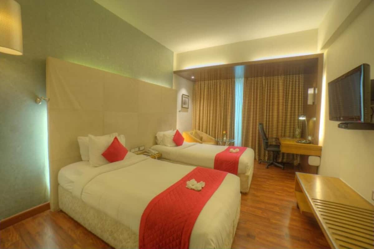 Quarto de hotel com duas camas, uma escrivaninha, uma cadeira, uma televisão e piso de madeira.