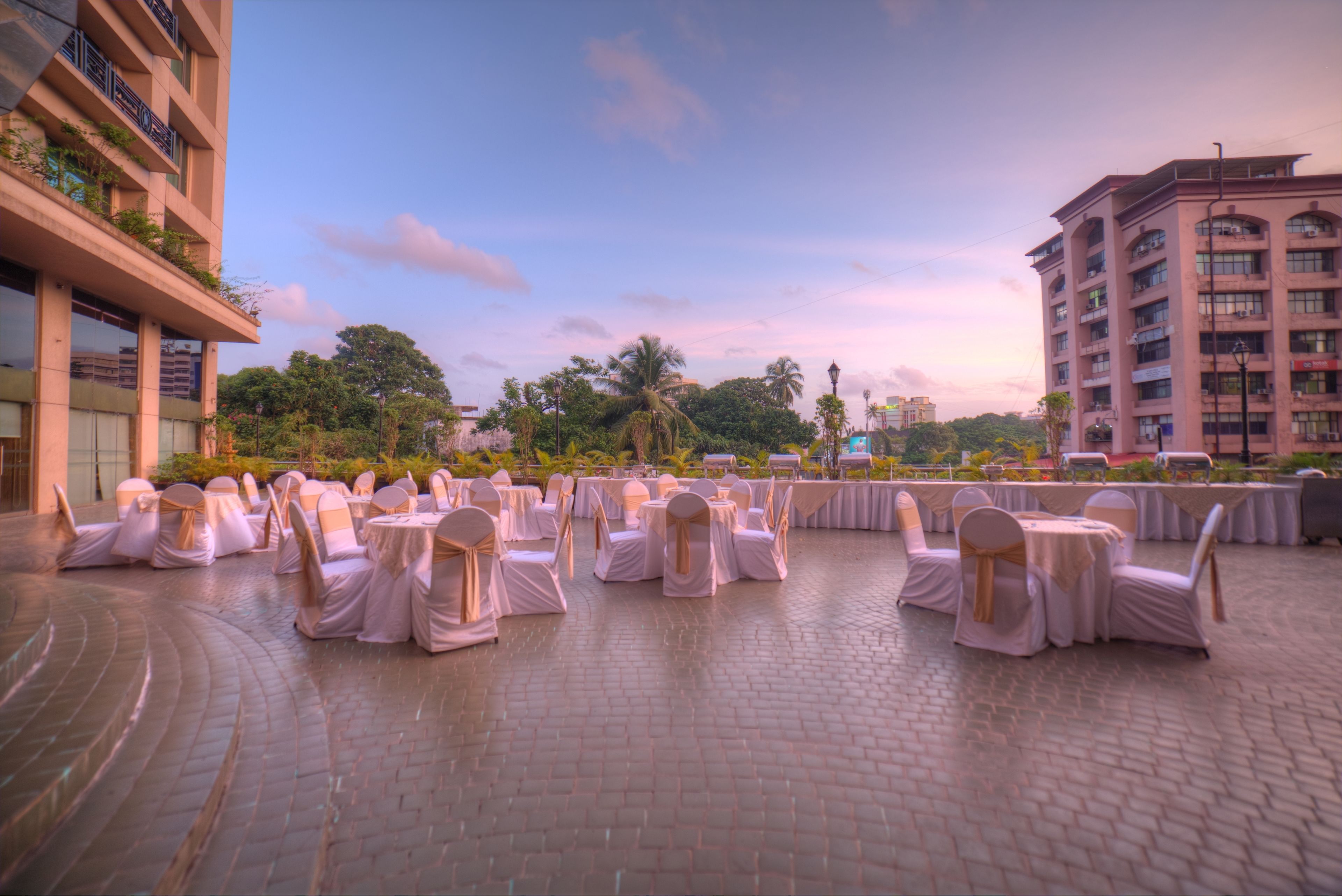 Um espaço para eventos ao ar livre, com mesas e cadeiras redondas, rodeado por edifícios e árvores.