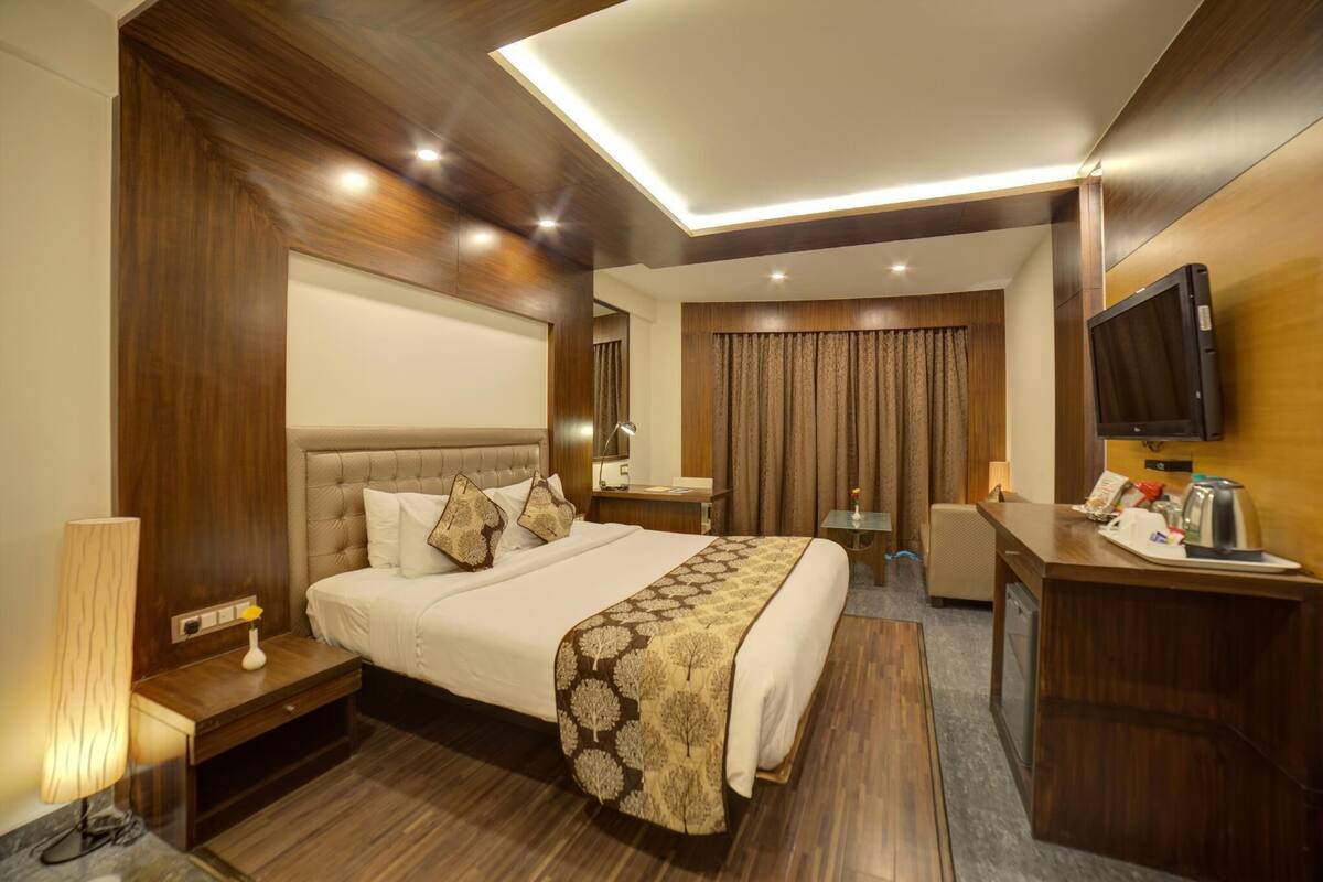 Quarto de hotel com cama, mesa de cabeceira, luminária, televisão e escrivaninha com cadeira.