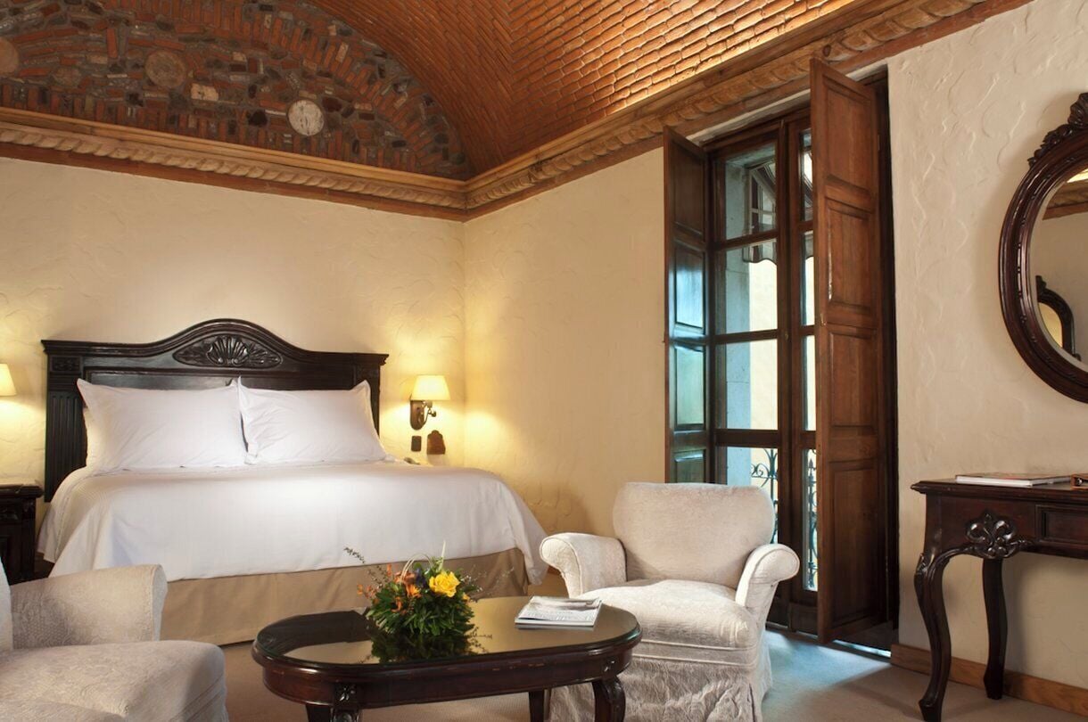 Quarto de hotel com uma cama grande, duas cadeiras, uma mesinha com um vaso e um espelho na parede.