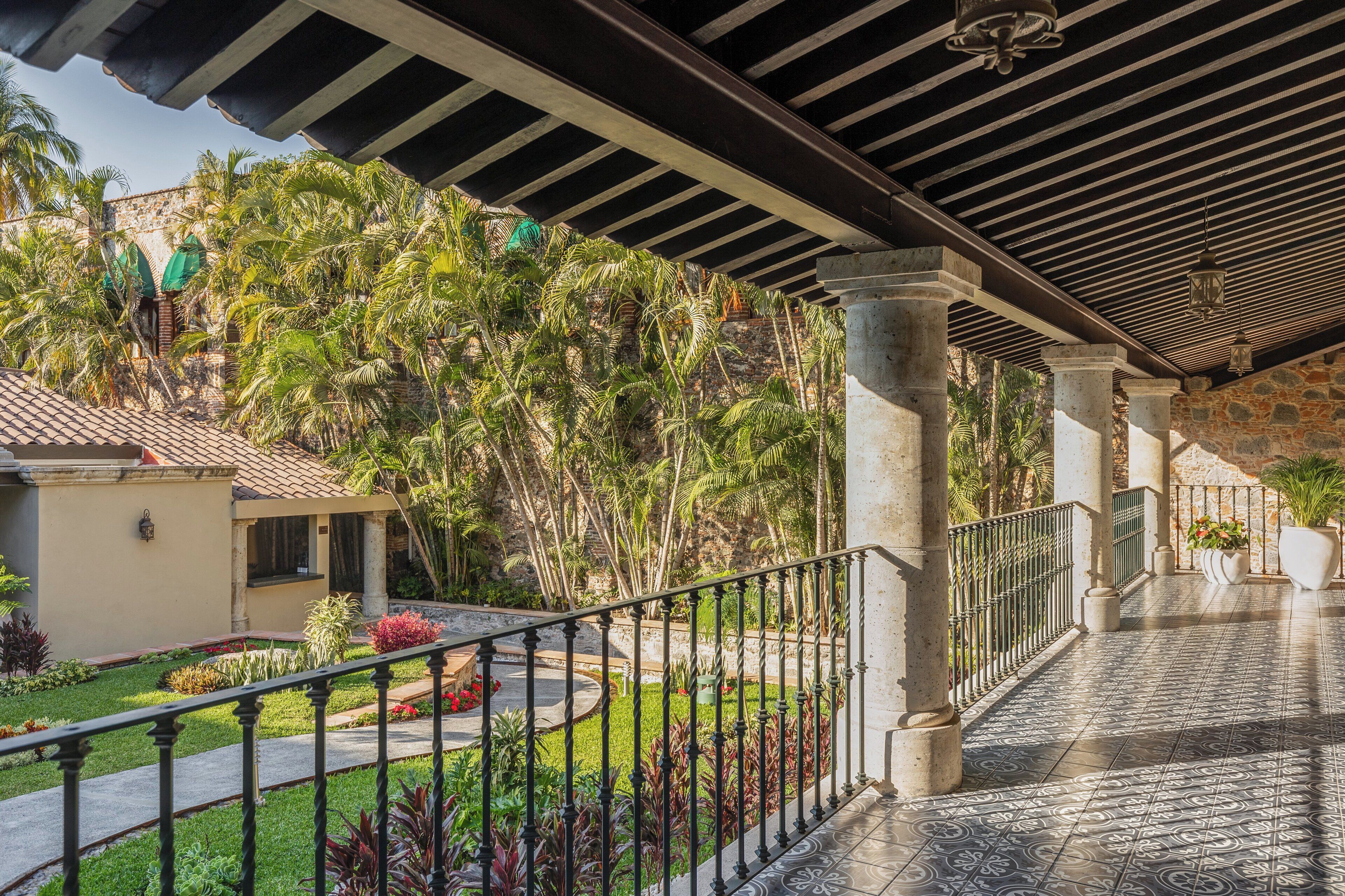Uma área externa coberta com piso de azulejos, corrimão e vista para um jardim com palmeiras e um edifício ao fundo.