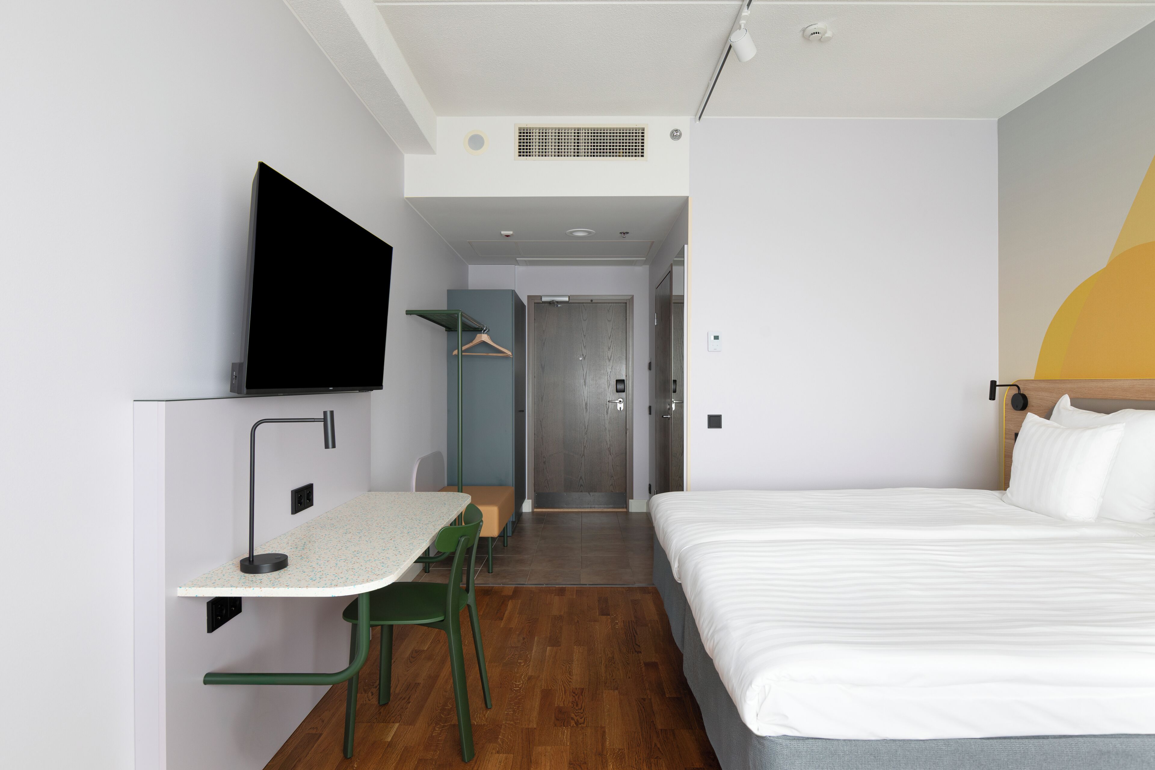 Quarto de hotel moderno com uma cama, televisão na parede, uma mesinha com cadeiras e chão de madeira.