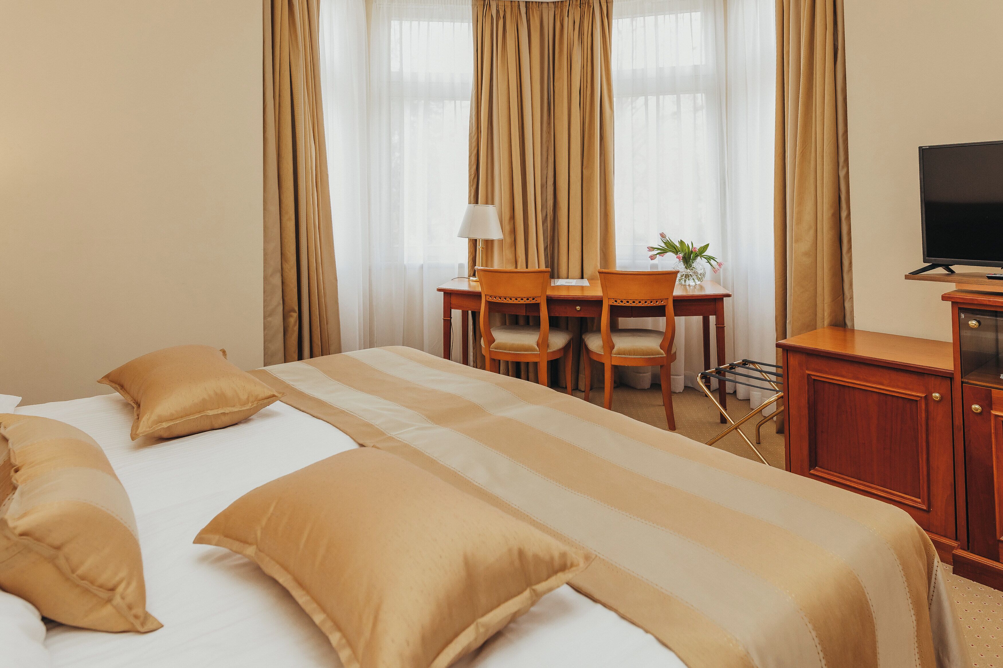 Quarto de hotel com cama, escrivaninha com cadeiras, televisão e janela com cortinas.