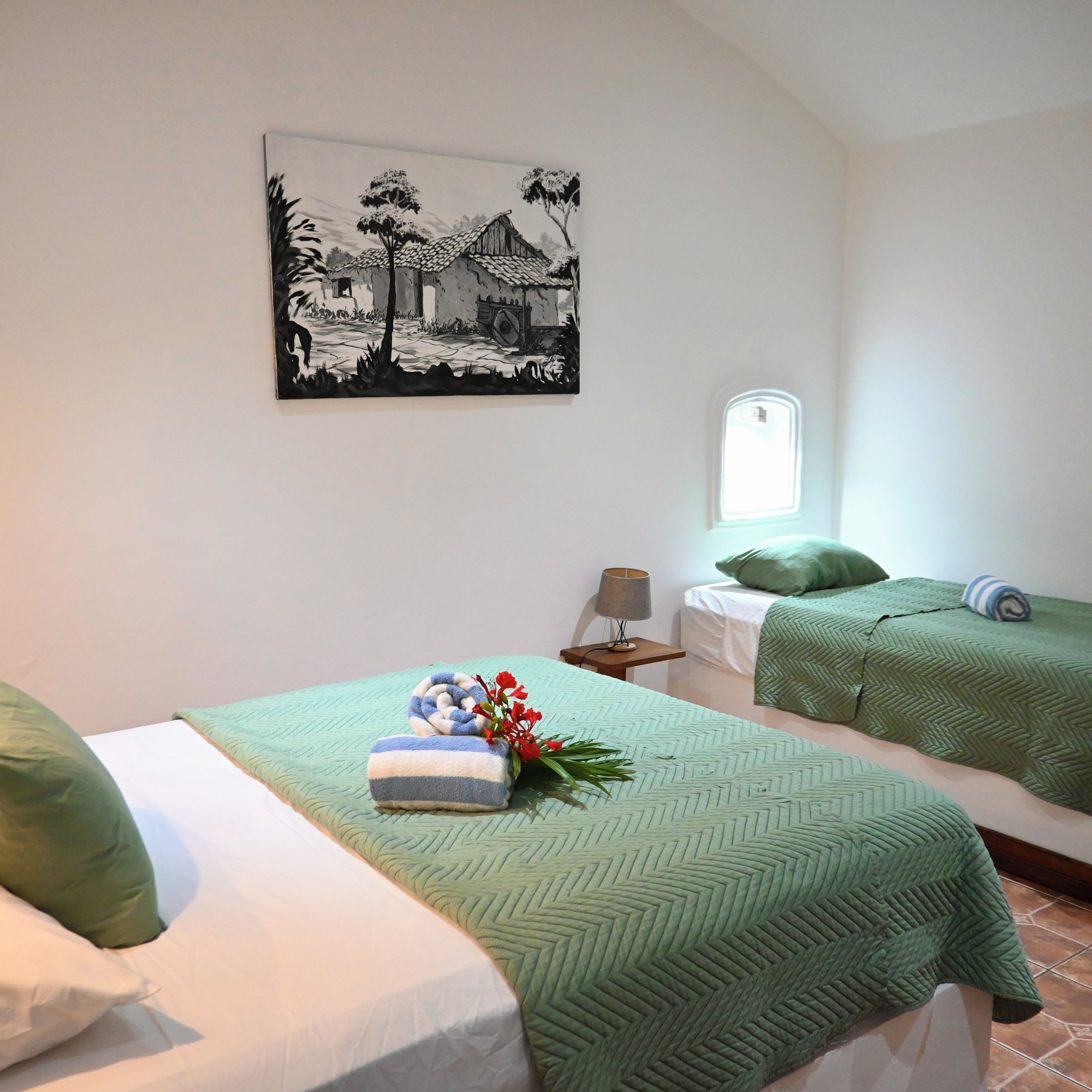 Quarto com duas camas, um quadro na parede, uma janela e uma mesa de cabeceira com abajur.
