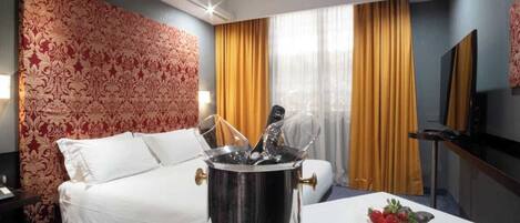 Quarto de hotel com cama, minibar com cubos de gelo e mesa com morangos e chocolate.