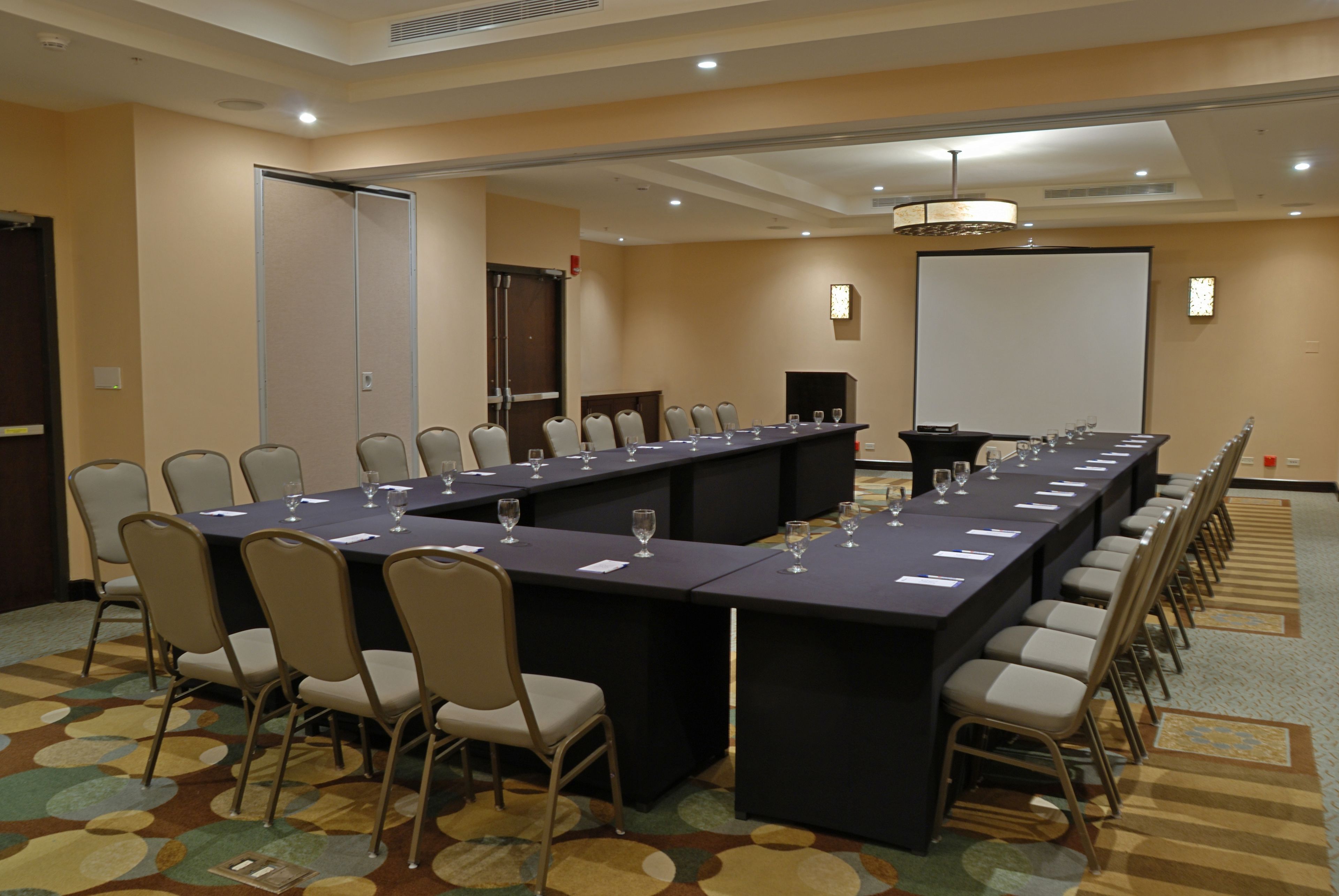 Sala de conferência com uma mesa comprida, cadeiras, tela de projetor e piso carpeteado.