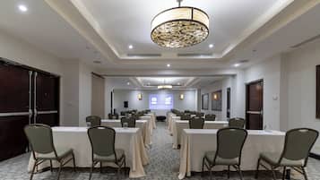 Uma sala de conferências com fileiras de mesas e cadeiras, uma tela grande na frente e uma luminária decorativa no teto.