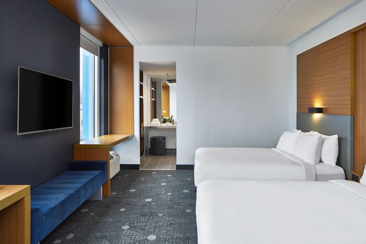 Quarto de hotel com duas camas, uma escrivaninha, um banco, televisão e cozinha compacta.