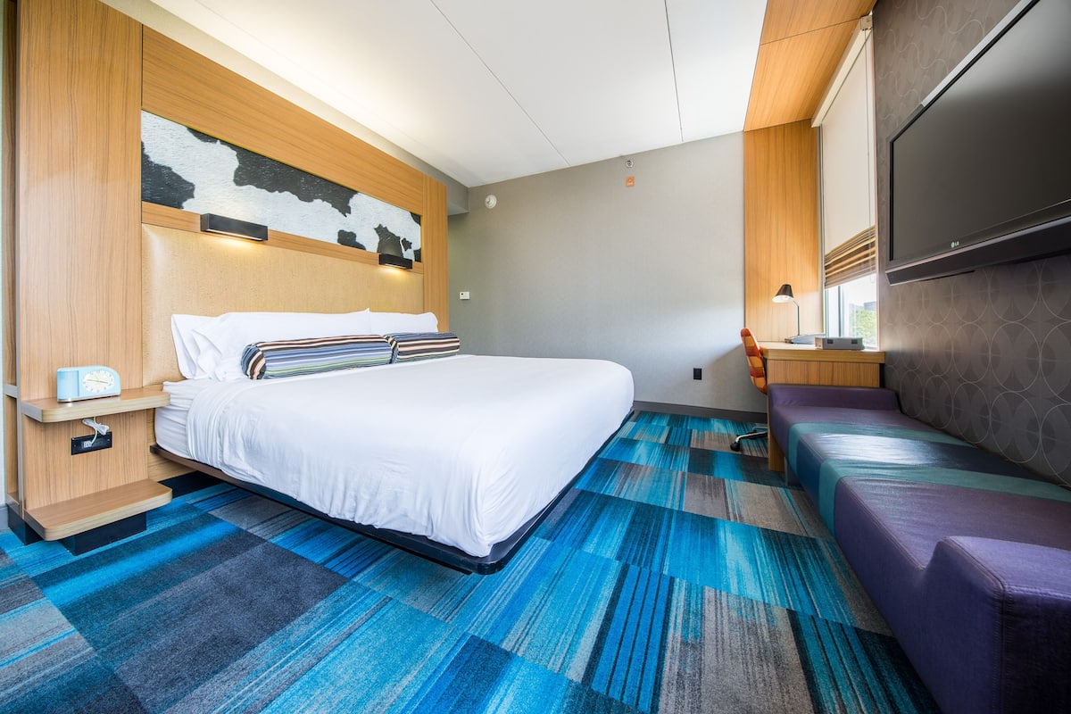 Quarto de hotel com uma cama grande, um sofá, uma televisão e uma cabeceira de madeira.
