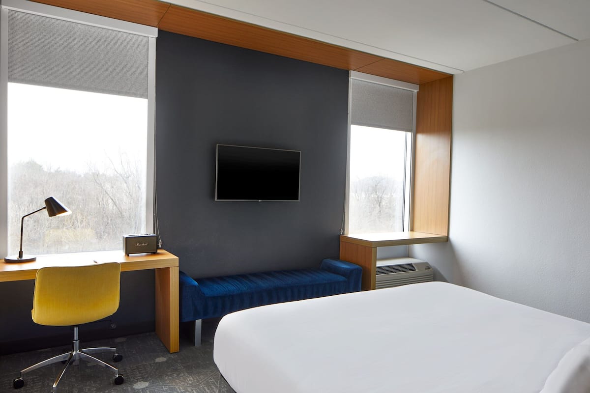 Quarto de hotel moderno com cama, escrivaninha, cadeira, televisão e um banco.