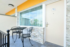 Uma varanda com mesa e cadeiras, uma janela com cortinas e uma porta com placa de número.