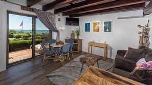 Uma sala de estar com piso de madeira, área de jantar com cadeiras de vime, um sofá e uma TV na parede.