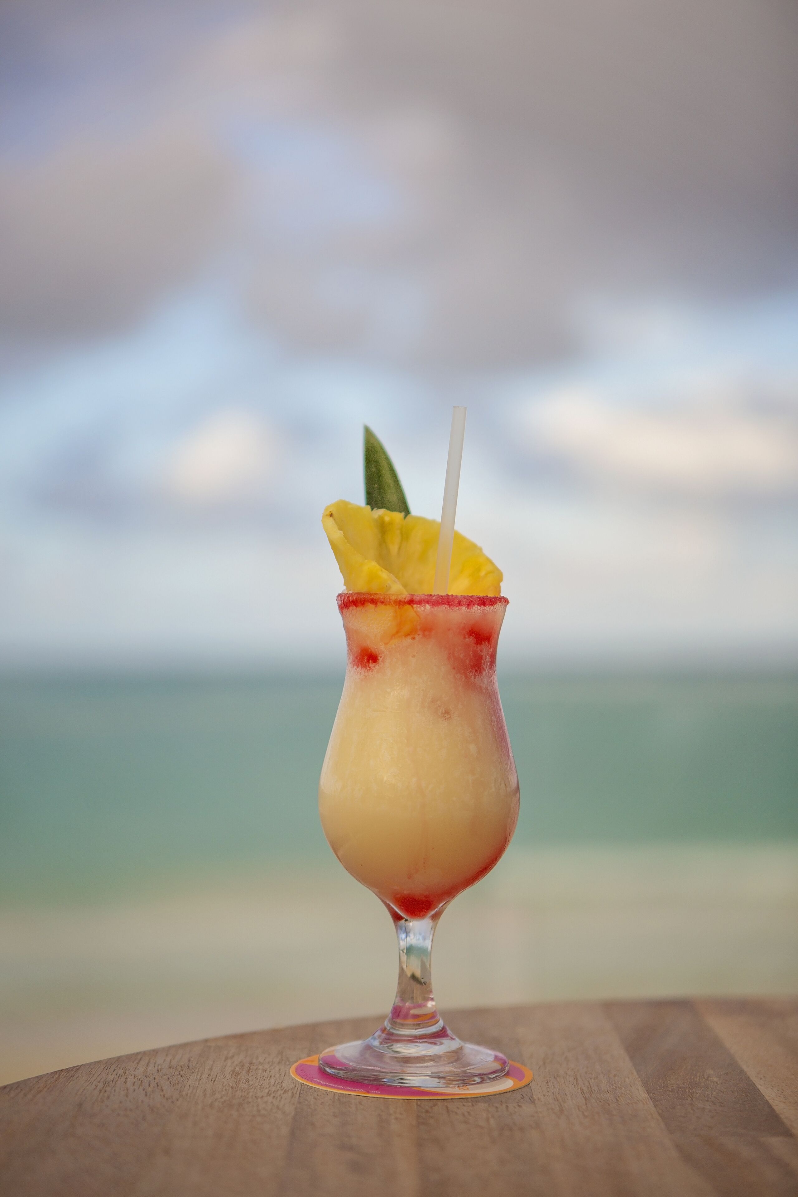 Um cocktail num copo com canudo, adornado com uma fatia de abacaxi, sobre uma mesa de madeira com oceano e céu desfocados ao fundo.