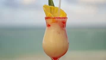 Um cocktail num copo com canudo, adornado com uma fatia de abacaxi, sobre uma mesa de madeira com oceano e céu desfocados ao fundo.