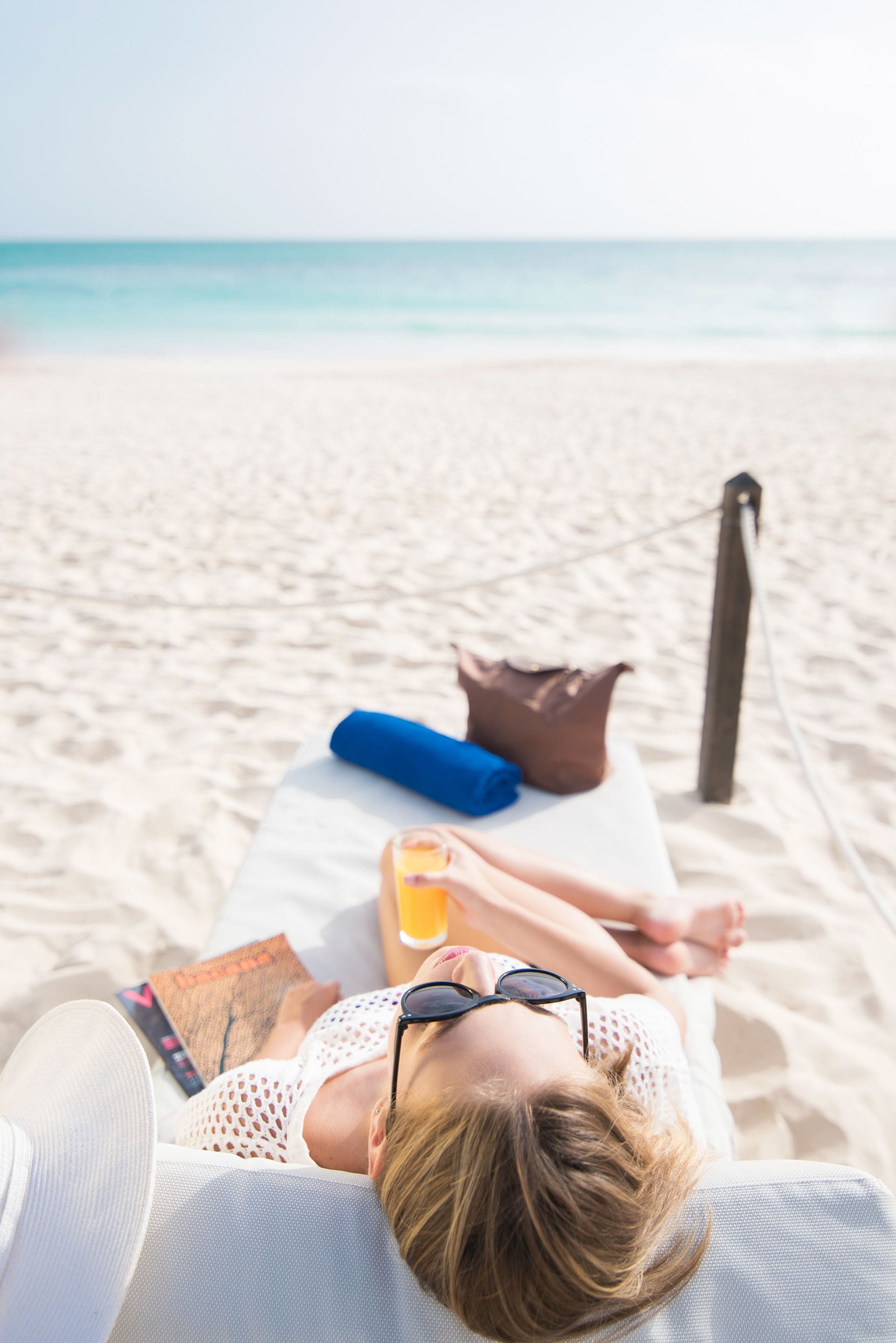 Uma pessoa deitada em uma toalha de praia, com um livro, um copo de bebida e uma bolsa.