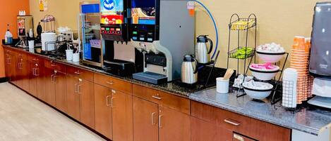 Um espaço de bebidas de serviço próprio, com várias máquinas de café, uma vitrine refrigerada e uma variedade de copos e recipientes descartáveis.