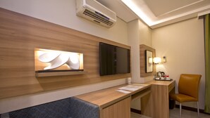 Quarto de hotel com escrivaninha de madeira, televisão fixada na parede, área de estar com uma cadeira e espelho.