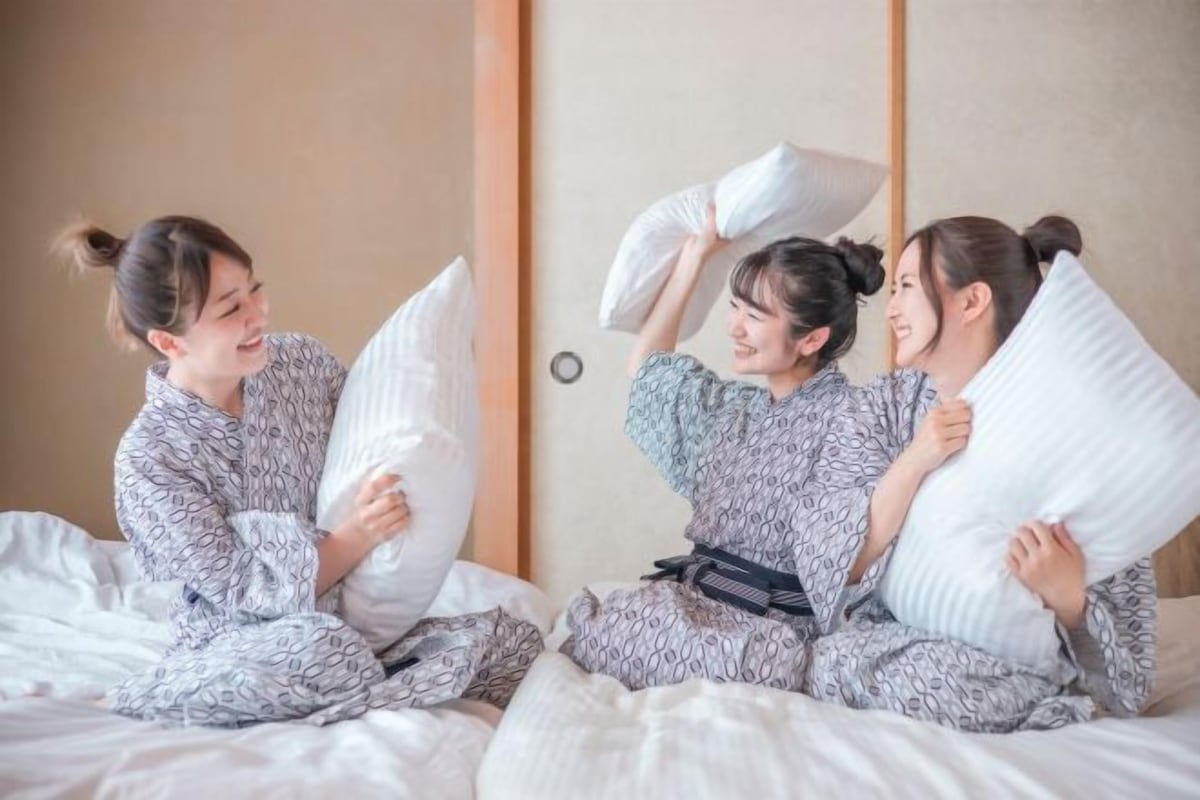 Três pessoas sentadas em uma cama com roupa de cama estampada, segurando travesseiros.