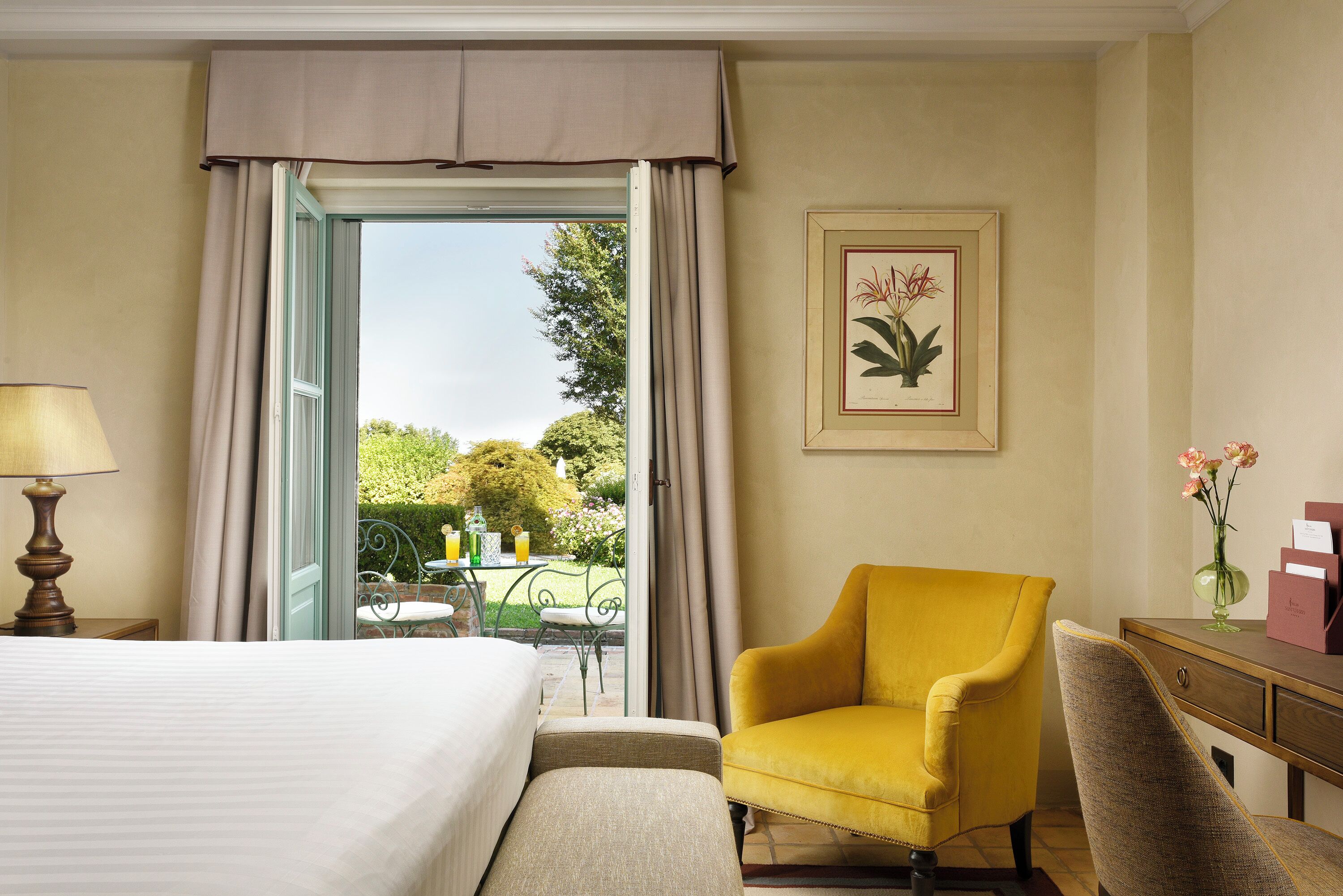 Quarto de hotel com uma cama, uma poltrona amarela, uma escrivaninha com um vaso de flores e uma ilustração botânica emoldurada na parede.