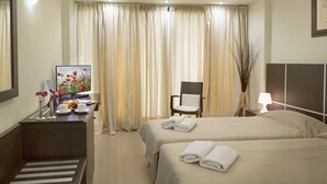 Quarto de hotel com duas camas, uma escrivaninha com computador e um vaso de flores, uma cadeira e uma janela grande com cortinas.
