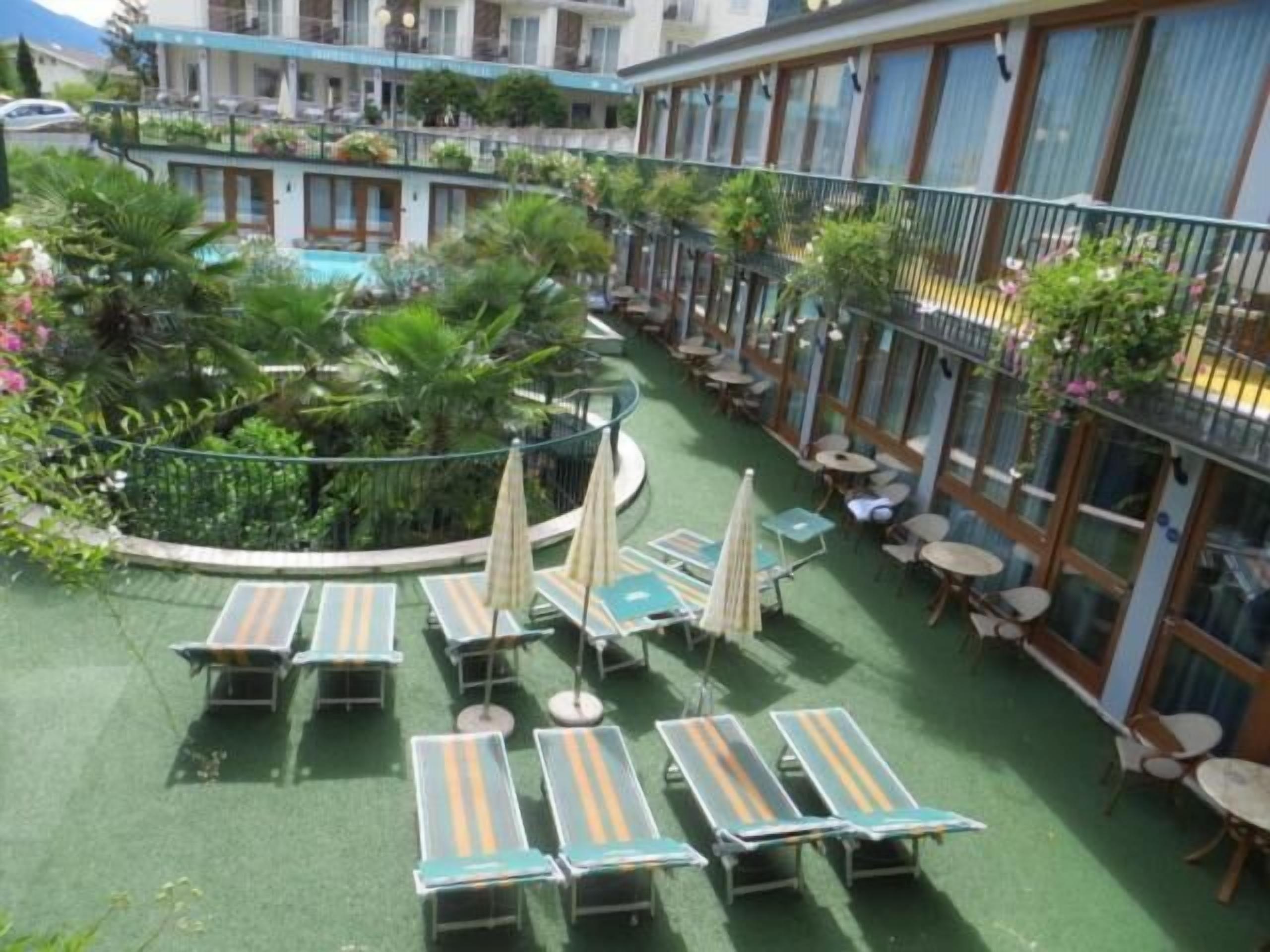 Pátio de hotel com espreguiçadeiras, pequenas mesas e guarda-sóis, rodeado por vegetação exuberante e uma piscina.
