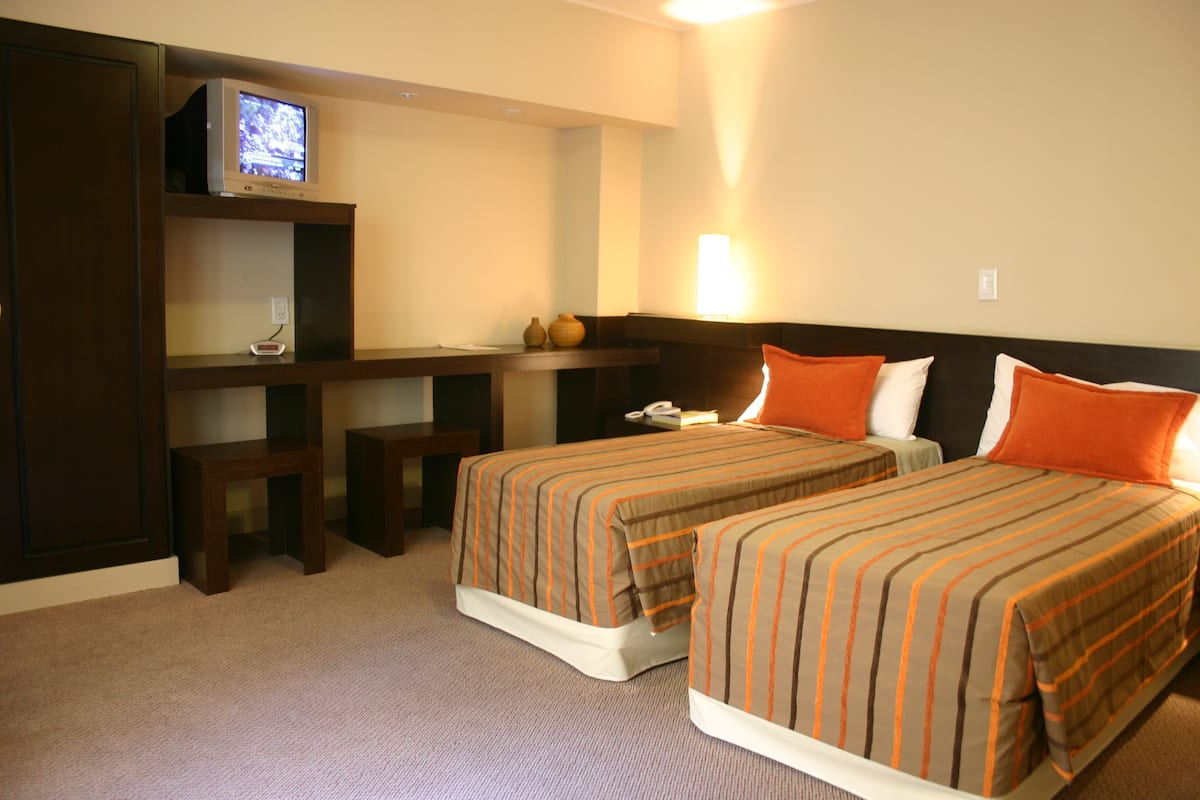 Quarto de hotel com duas camas, uma escrivaninha, televisão e mesa de cabeceira.