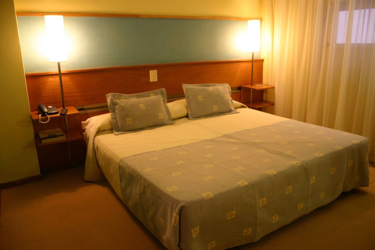 Quarto de hotel com uma cama grande, duas luminárias de cabeceira, mesa de cabeceira com telefone e uma janela com cortinas.