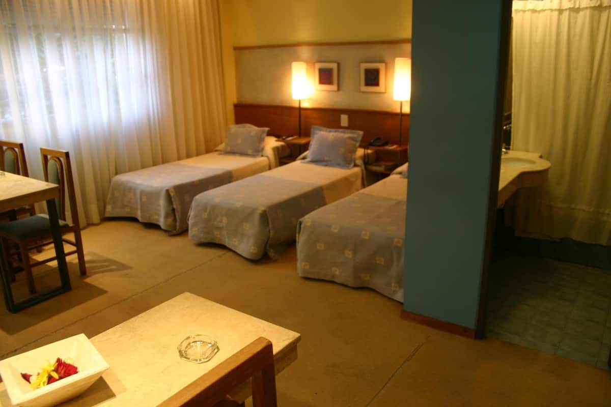 Quarto de hotel com duas camas de solteiro, uma mesinha com uma cesta de frutas, uma cadeira e uma mesa de cabeceira com abajur.