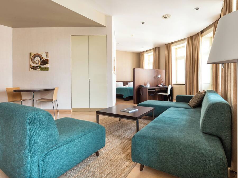 Quarto de hotel moderno com sofás verde-água, área de refeições, escrivaninha e uma janela grande com cortinas.
