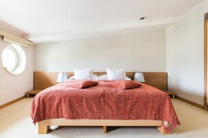 Cama bem arrumada com colcha xadrez vermelha e almofadas brancas, um relógio de parede redondo e uma mesa de cabeceira com abajur.
