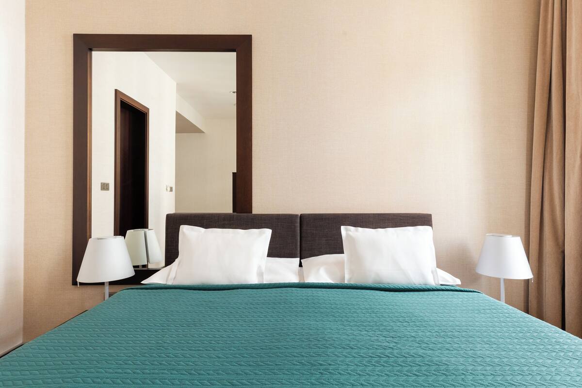 Quarto de hotel com cama grande, duas luminárias de cabeceira, um espelho e vista para o corredor.